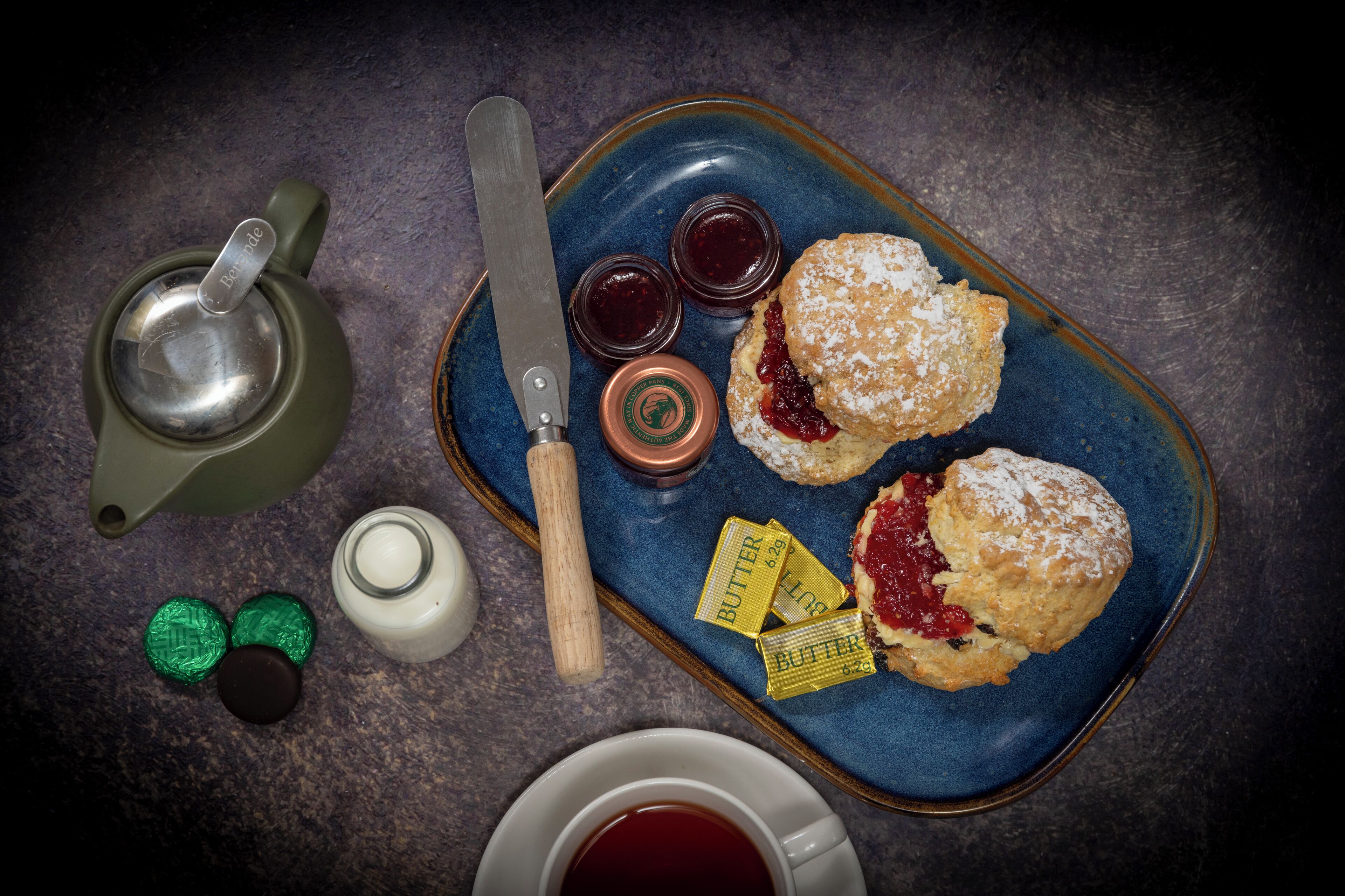 Uma bandeja com dois scones, dois potes de geleia, uma faca de manteiga, um bule de chá e uma xícara de chá.