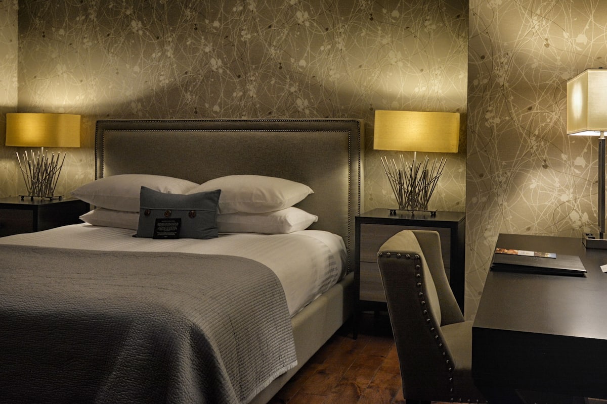 Quarto de hotel com uma cama grande, duas mesas de cabeceira com luminárias, uma cadeira e uma escrivaninha com luminária.