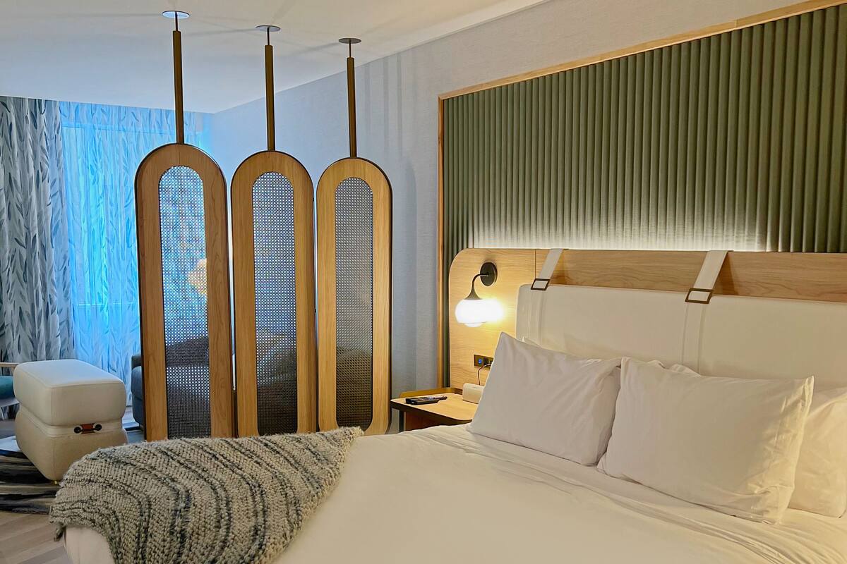 Quarto de hotel com uma cama grande, mesa de cabeceira com abajur, cadeira e janela com persianas.