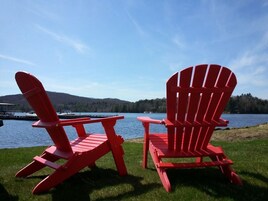 Duas cadeiras Adirondack vermelhas à beira de um lago, com um cais e barcos ao fundo.