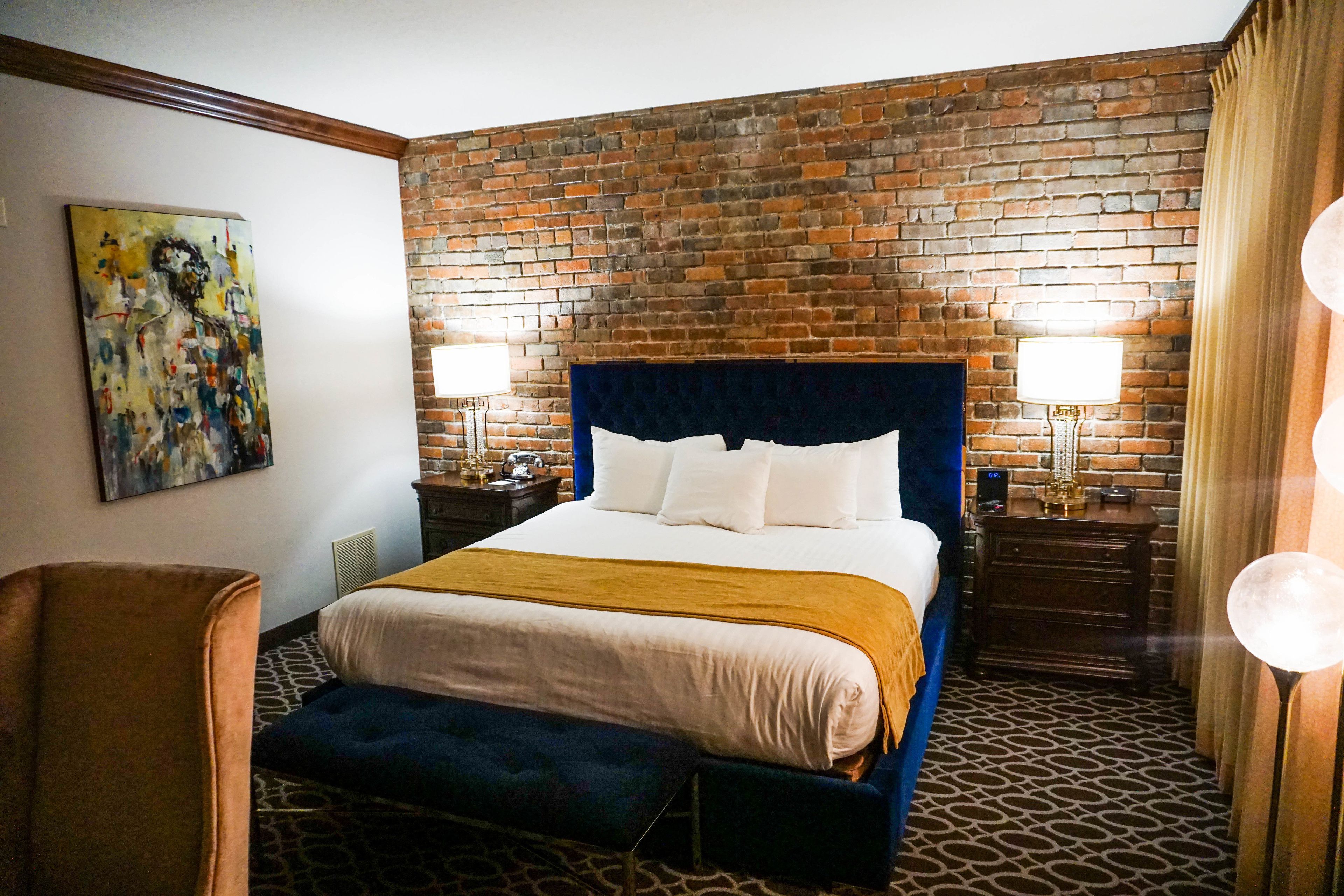 Quarto de hotel com parede de tijolos, uma cama grande com roupa de cama branca e azul, duas mesas de cabeceira com abajures, e um quadro na parede.