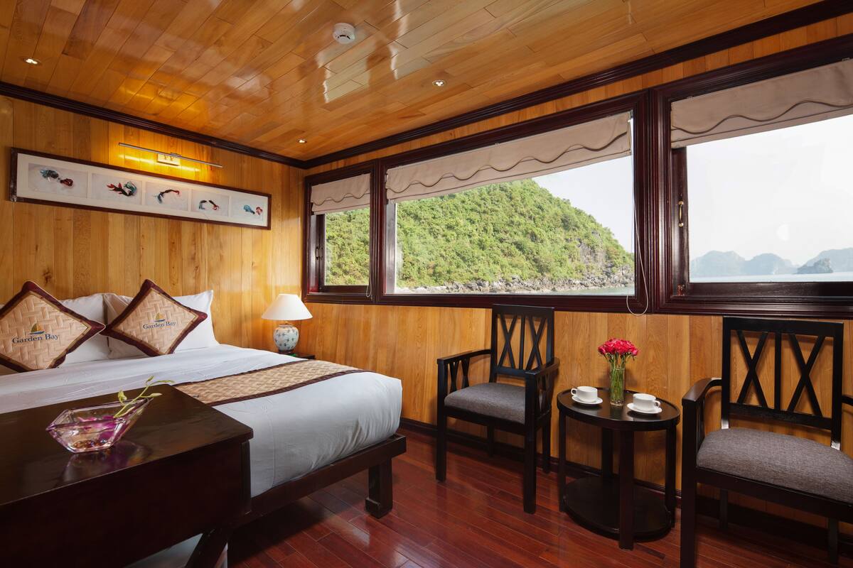 Quarto de hotel com teto de madeira, uma cama com almofadas, uma mesa de cabeceira com abajur, duas cadeiras, uma pequena mesa com xícaras e um vaso, e uma janela com vista para uma montanha.