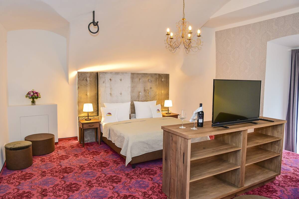 Quarto de hotel com uma cama grande, um móvel de televisão de madeira, um candelabro e um tapete vermelho estampado.