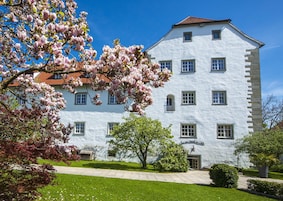 Um edifício branco com várias janelas e telhado vermelho, rodeado por relva verde e árvores com flores.