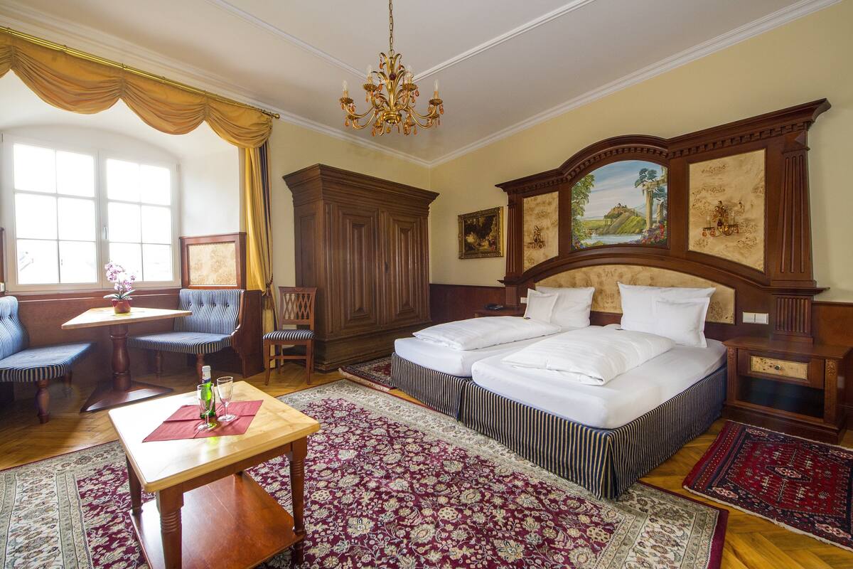 Quarto de hotel com cama de casal, cabeceira de madeira, lustre, área de refeições com mesa e cadeiras, e uma janela grande com cortinas.