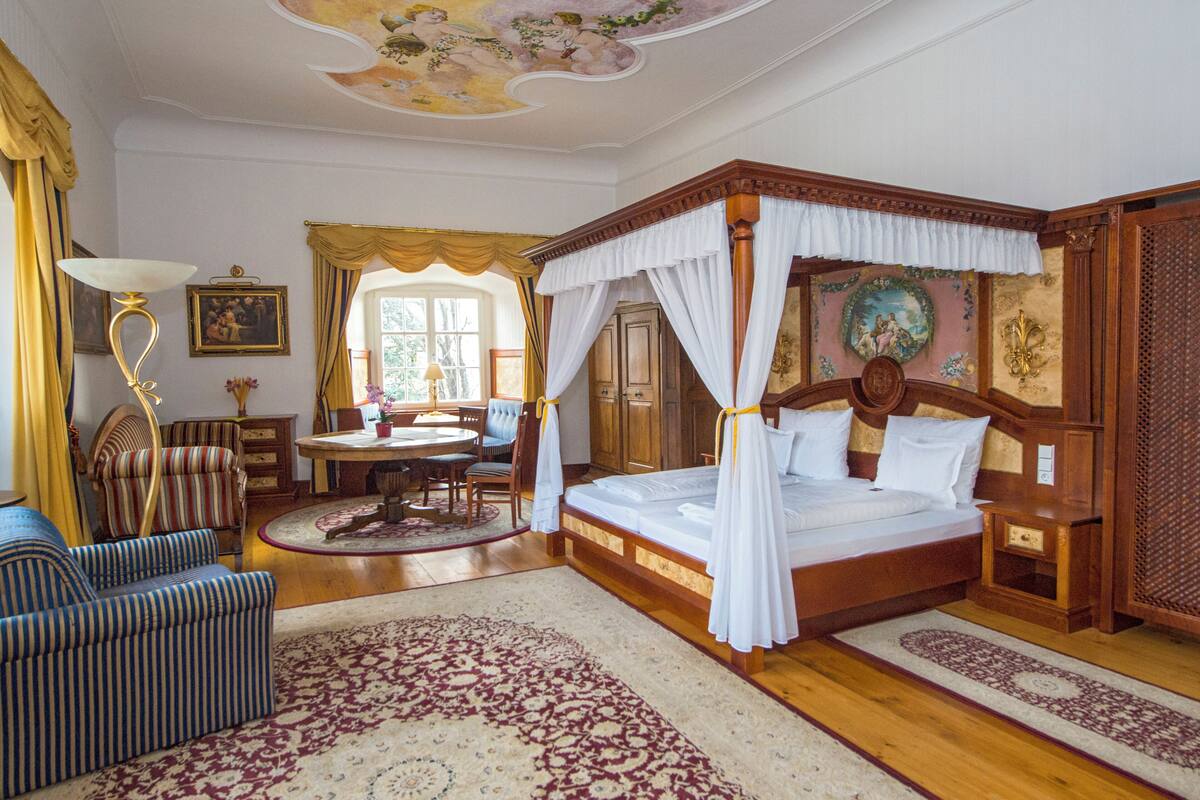 Quarto espaçoso com cama de dossel, área de estar com mesa e cadeiras, e uma grande janela que oferece luz natural.