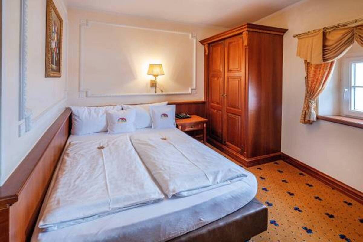 Quarto de hotel com cama, mesa de cabeceira, guarda-roupa de madeira, candeeiro fixado na parede e janela com cortinas.