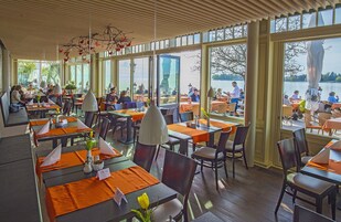 Restaurante com mesas preparadas para refeições, amplas janelas com vista para um lago e teto de madeira.