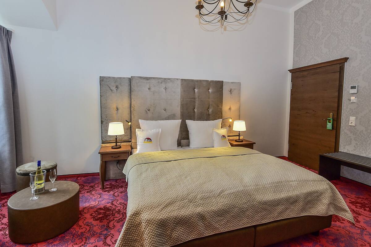 Quarto de hotel com uma cama grande, duas mesas de cabeceira com luminárias, um guarda-roupa de madeira e uma mesa lateral redonda com uma garrafa e copos.