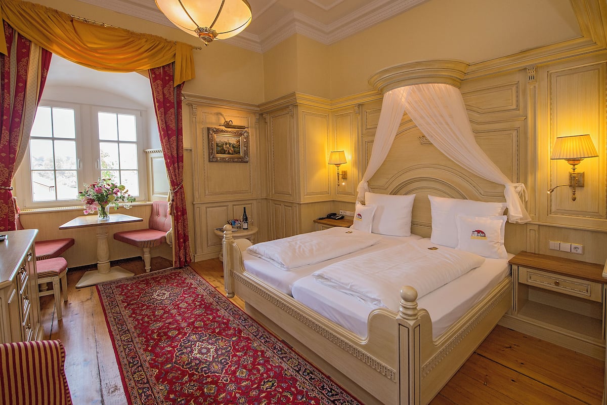 Quarto com cama de dossel, área de estar com mesa e cadeiras, e uma grande janela com cortinas.