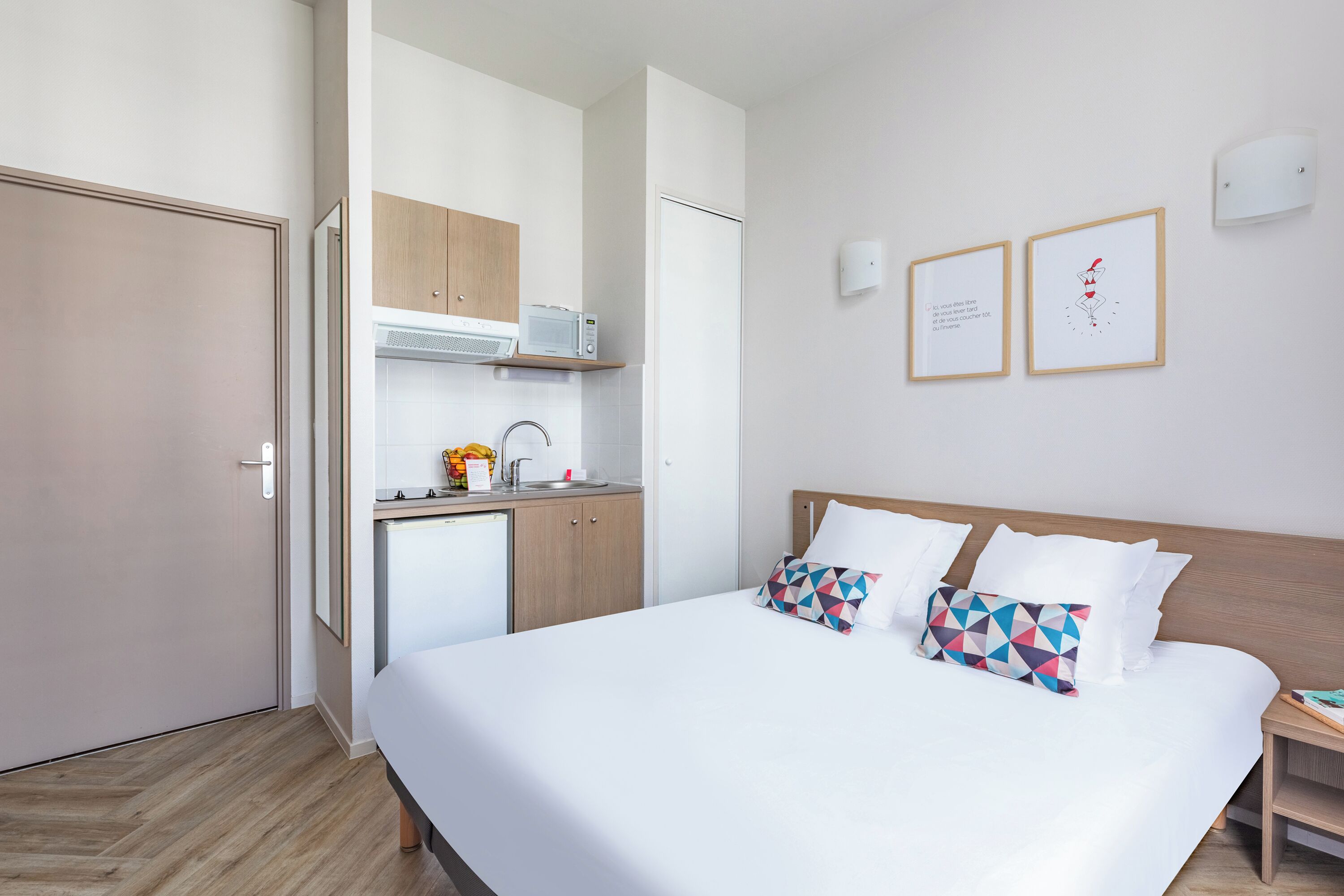 Quarto de hotel com cama, uma pequena copa, uma porta e duas imagens emolduradas na parede.