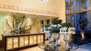 Um hall com lustres, mesa de centro de vidro, um detalhe decorativo na parede e uma grande janela com vista para palmeiras.