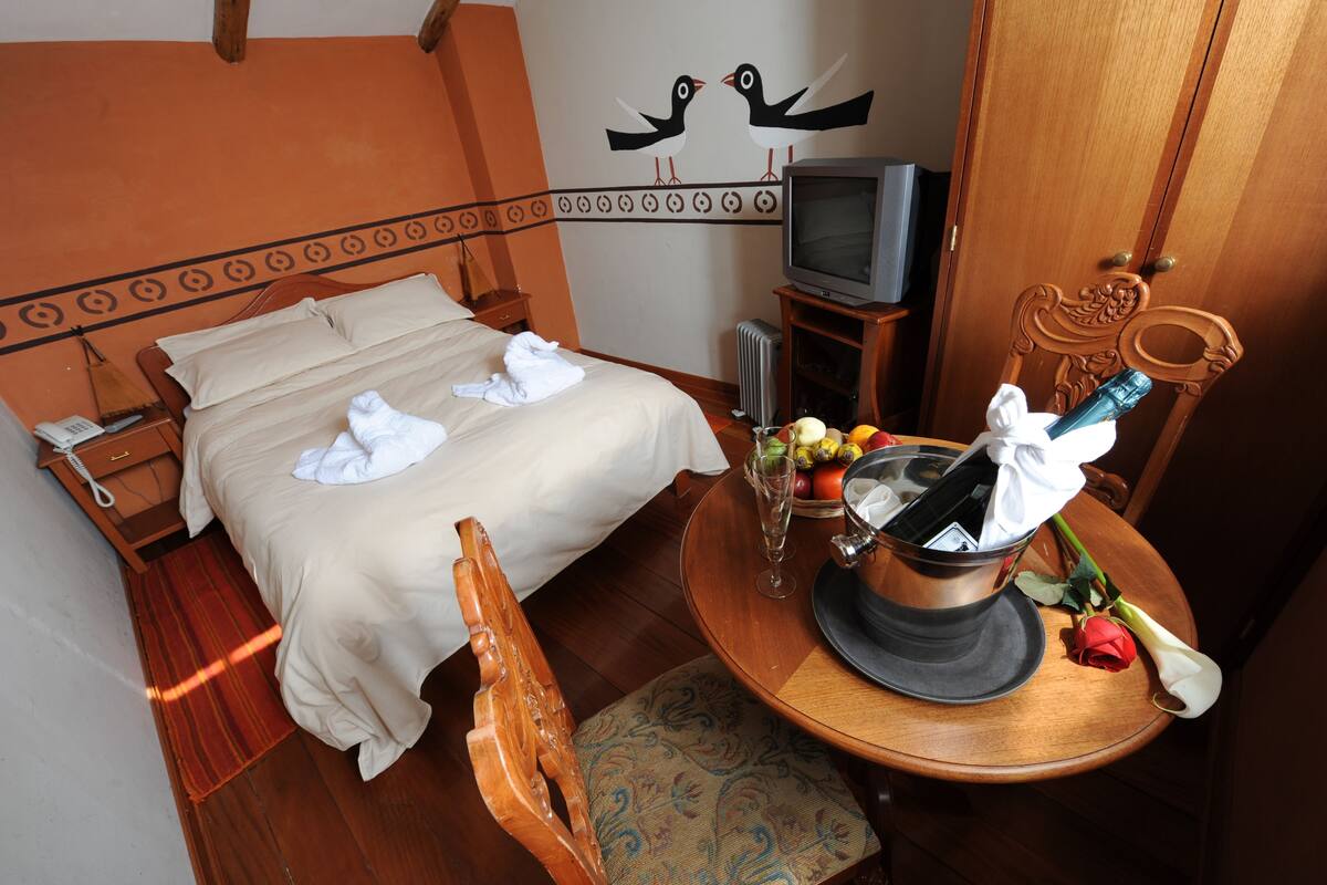 Quarto de hotel com cama, televisão, mesa de jantar com um vaso de flores e uma parede decorativa com pássaros.
