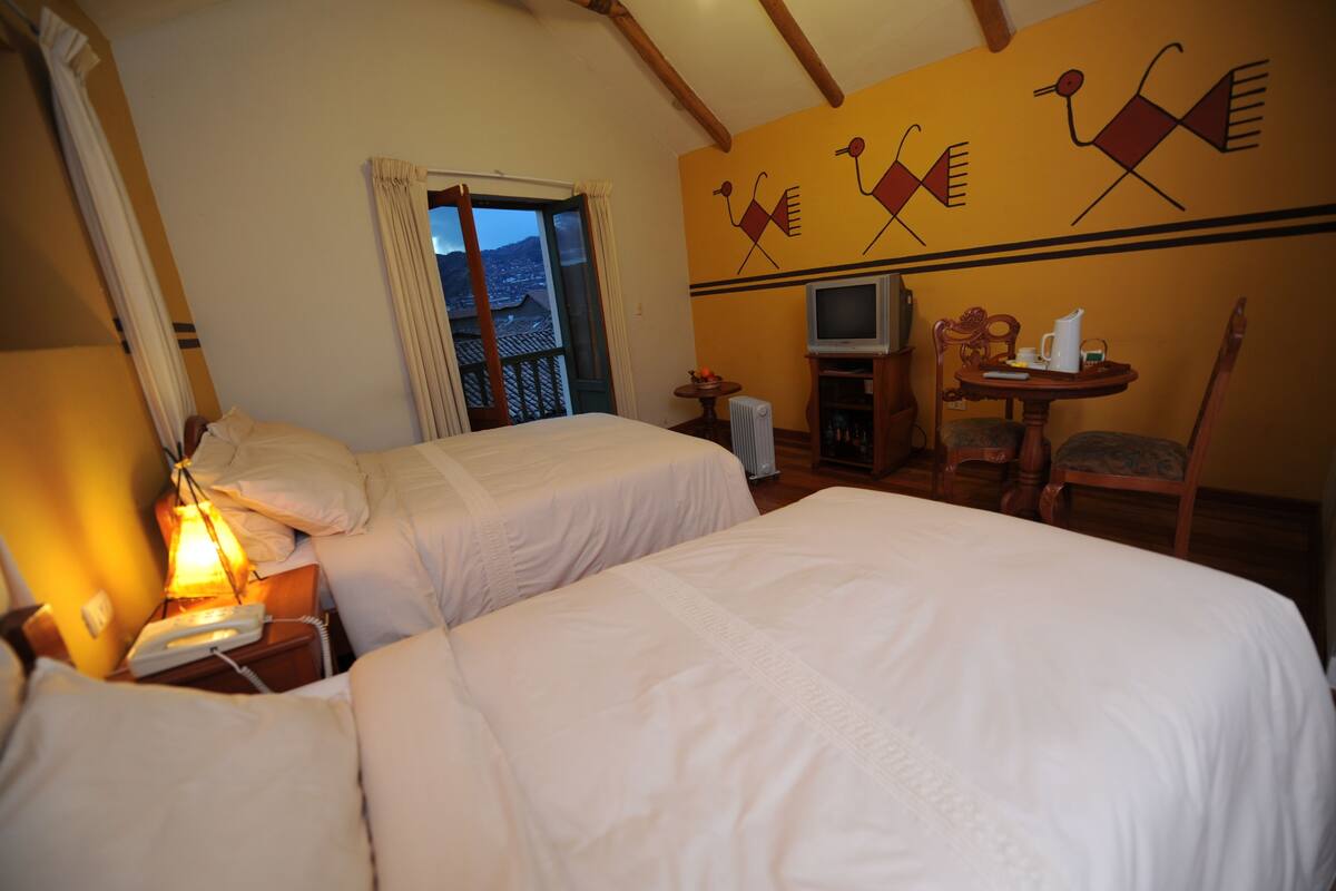 Quarto de hotel com duas camas, televisão, mesa pequena com cadeira e janela com cortinas.