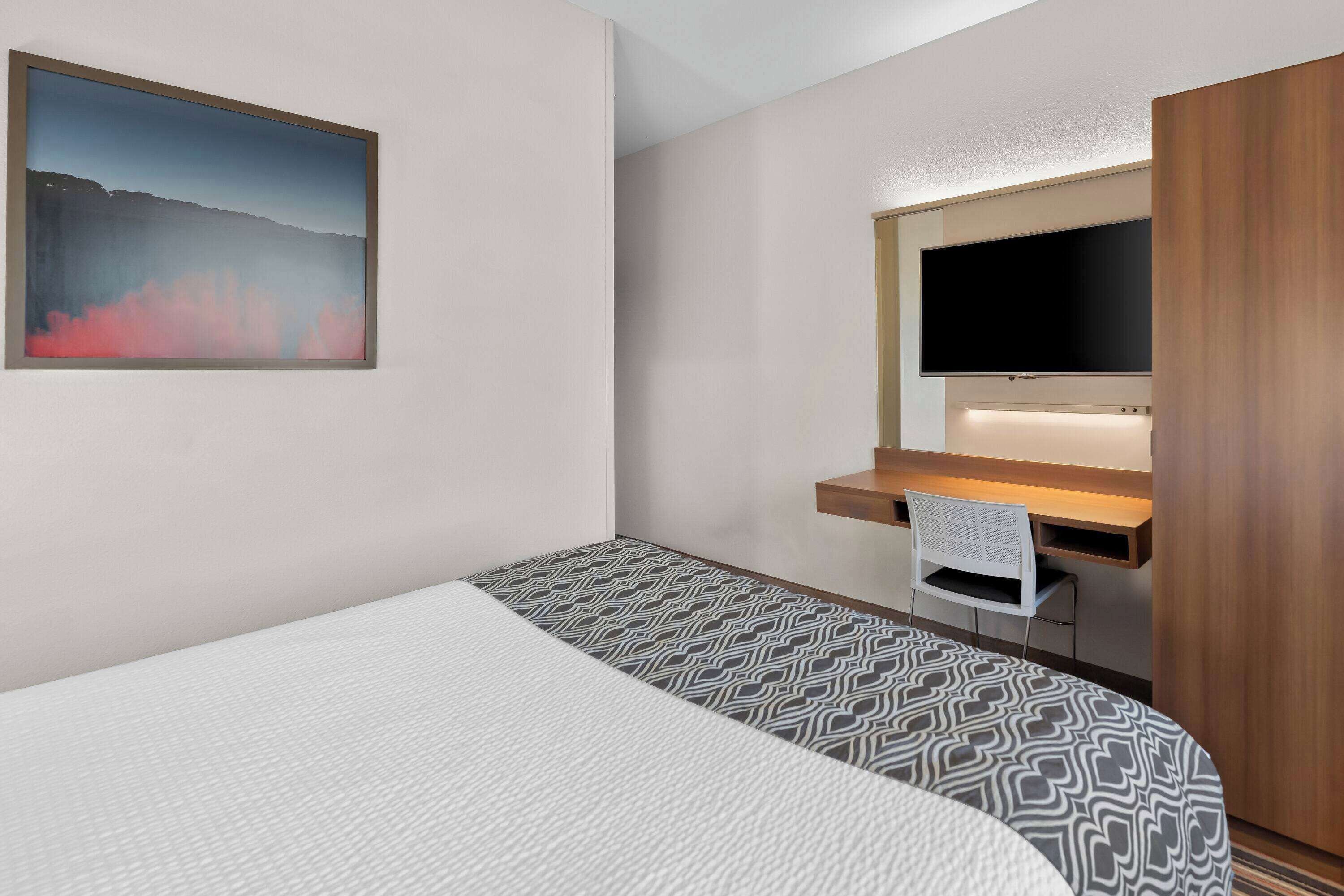 Quarto de hotel com uma cama, uma escrivaninha com cadeira, televisão na parede e um guarda-roupa.