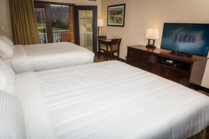 Quarto de hotel com duas camas, uma escrivaninha, televisão e janela com vista para árvores.