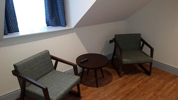 Quarto pequeno com duas cadeiras e uma mesa redonda, janela com cortinas e piso de madeira.