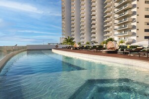 Piscina com espreguiçadeiras e guarda-sóis, cercada por um edifício alto com varandas.