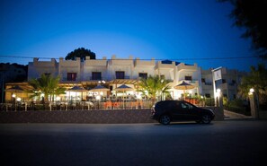 Hotel com área de estar externa e guarda-sóis, um carro estacionado na frente e um céu claro à noite.