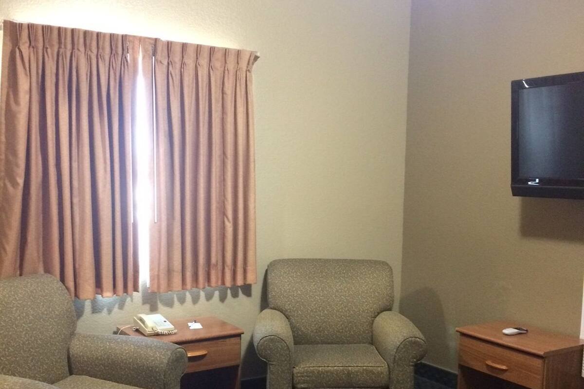 Quarto de hotel com duas poltronas, uma mesa de centro, televisão de tela plana, escrivaninha com telefone e janela com cortinas.