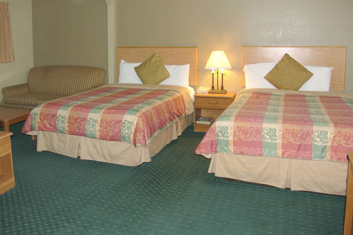 Quarto de hotel com duas camas de solteiro, uma cadeira, mesa de cabeceira com abajur e uma pequena mesa.