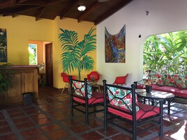 Um quarto com soalho em azulejos, um banco de madeira, cadeiras vermelhas, uma pequena mesa e uma porta que dá para outro espaço.
