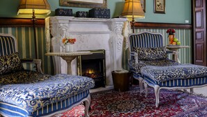 Quarto tradicional com lareira, dois poltronas estampadas, uma mesinha de centro com um vaso e um tapete estampado.
