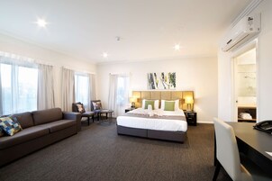 Quarto de hotel com uma cama grande, um sofá, uma escrivaninha e uma cadeira. Há uma janela com cortinas, um ar condicionado fixado na parede e um quadro na parede.