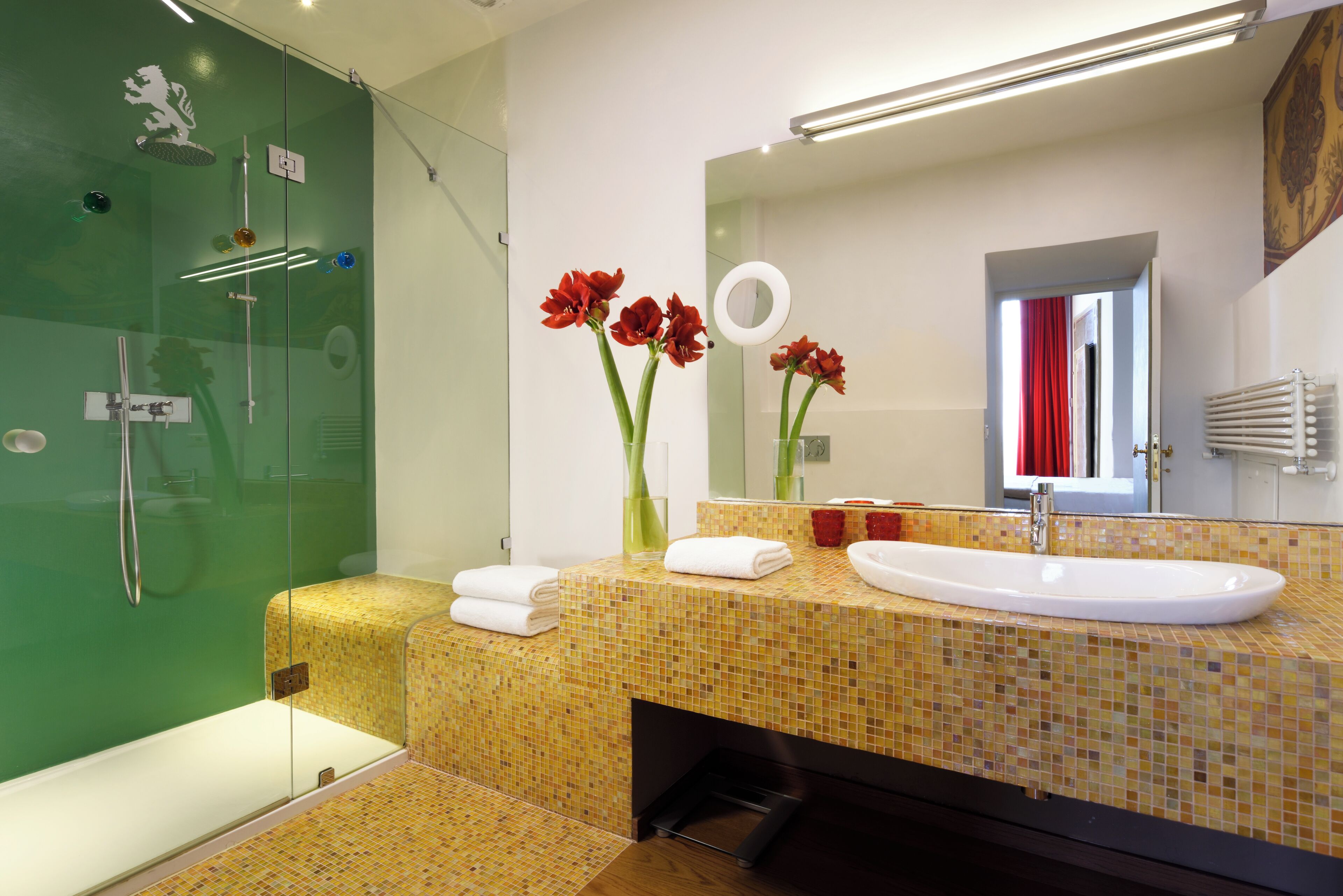 Um banheiro moderno com box de vidro, uma pia grande com espelho e flores vermelhas decorativas.