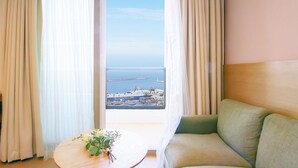 Quarto de hotel com vista para um porto e para a água. Há um sofá verde e uma mesinha com um arranjo de flores.