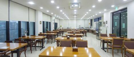 Uma ampla área de refeições com várias mesas e cadeiras, uma cozinha ao fundo e grandes janelas em um dos lados.