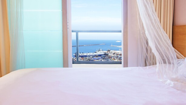 Quarto de hotel com uma cama e vista para o mar, com um navio chamado SUNWOO LINE ao fundo.