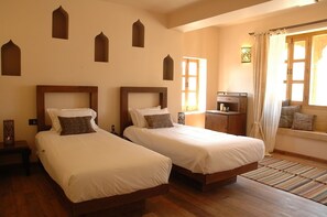 Quarto de hotel com duas camas de solteiro, cabeceiras de madeira, uma mesinha com abajur, janela com cortinas e vista para o exterior.