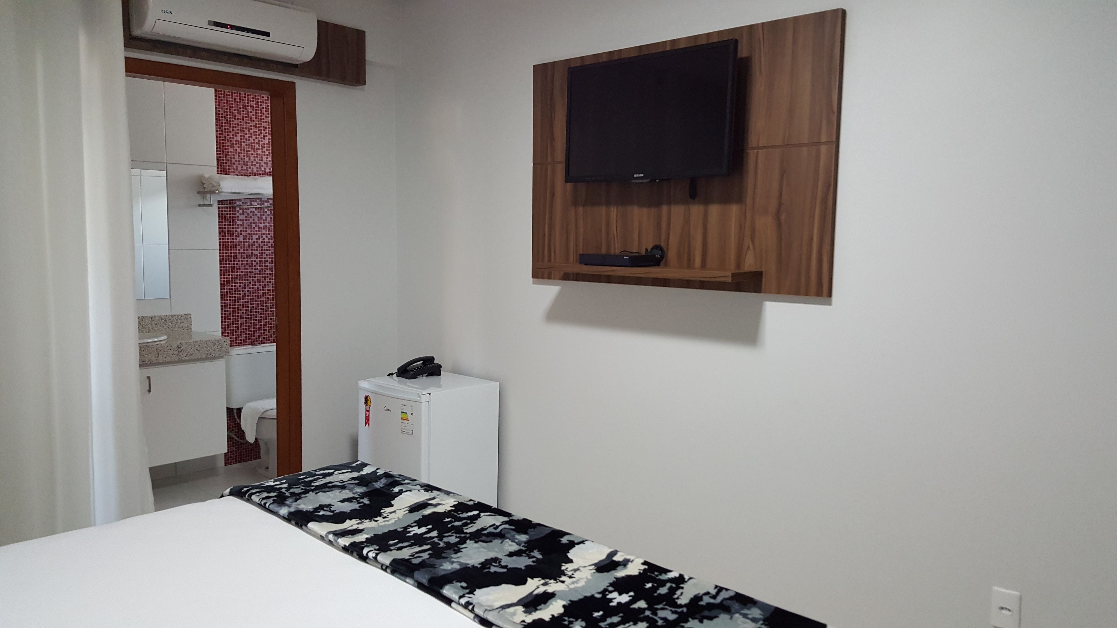 Quarto com cama, TV fixada em um painel de madeira, eletrodoméstico pequeno branco e banheiro visível por uma porta aberta.