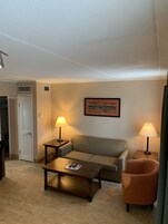 Uma sala de estar com um sofá, uma mesa de centro de madeira, duas luminárias, um quadro na parede e uma porta com persianas.