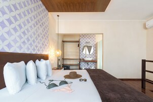 Quarto de hotel com cama, almofadas, chapéu, óculos de sol e mesa de cabeceira com abajur.
