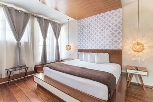 Quarto de hotel moderno com uma cama grande, piso de madeira, painel de cabeceira com padrão geométrico na parede e duas luminárias pendentes.