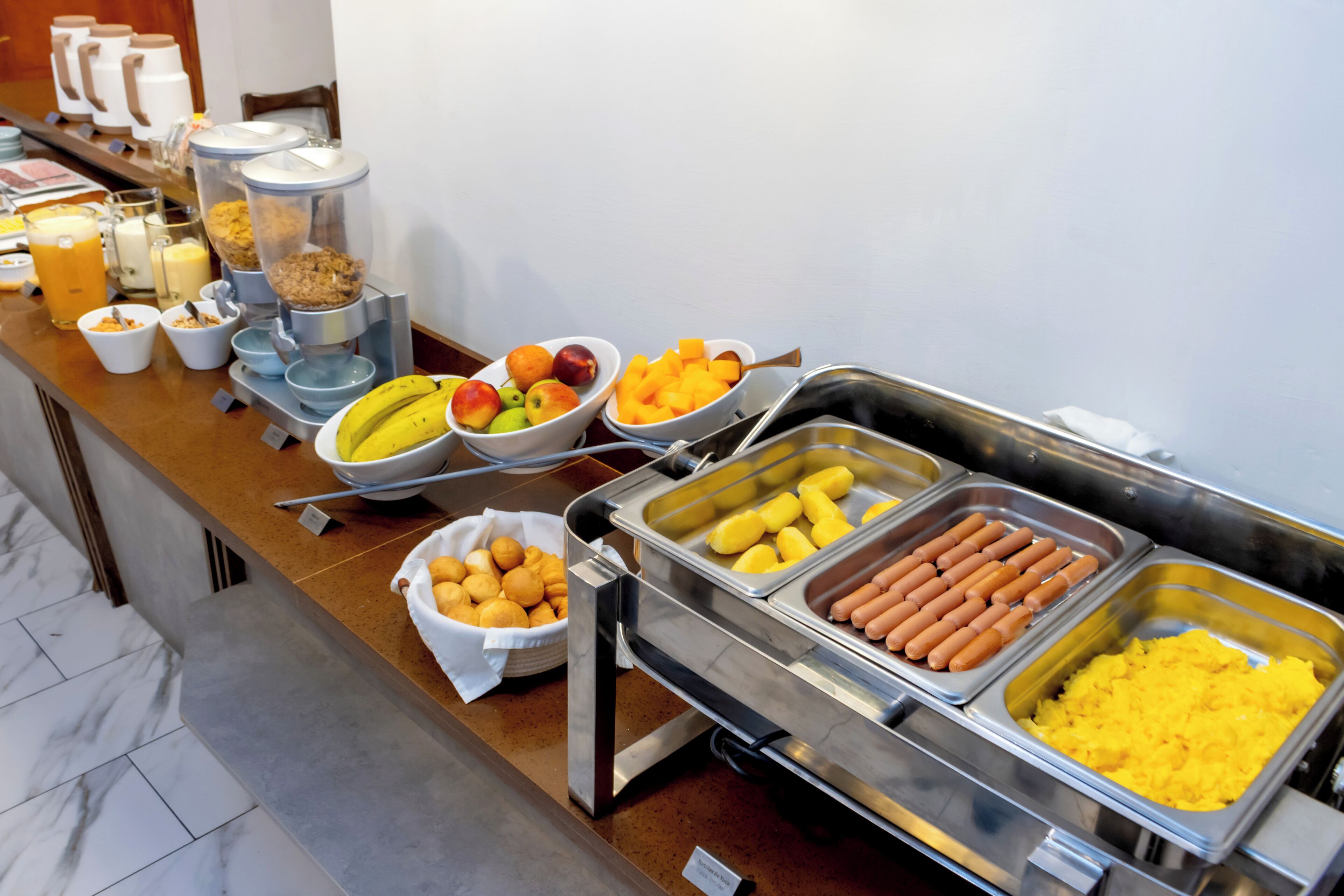 Buffet com diversos alimentos, incluindo frutas, cachorros-quentes e batatas.