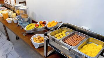 Buffet com diversos alimentos, incluindo frutas, cachorros-quentes e batatas.