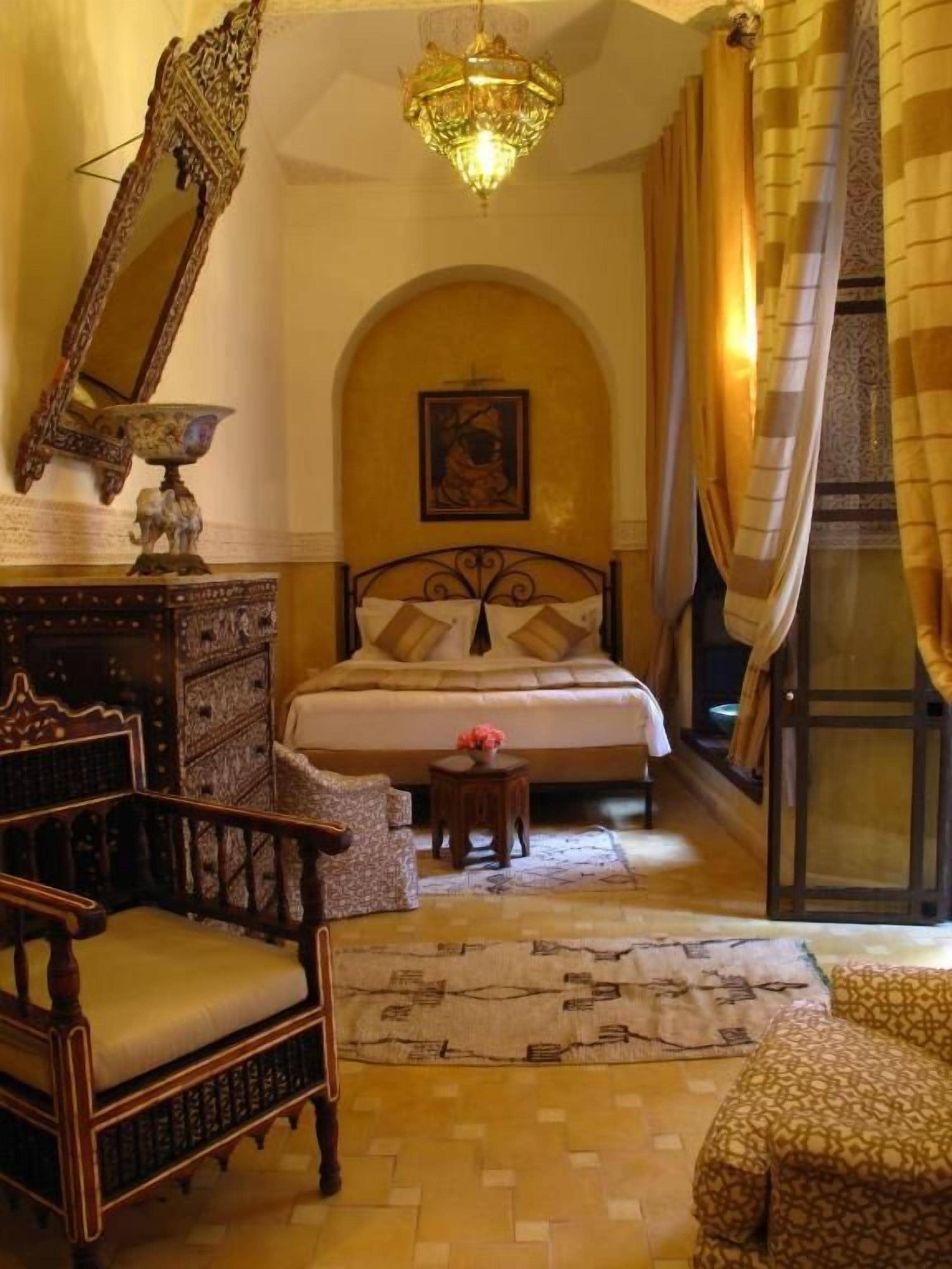 Um quarto com uma cama, uma cadeira, uma mesinha, um espelho e um candelabro.