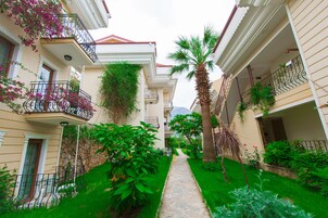 Um caminho estreito ladeado por vegetação exuberante e uma palmeira, levando a um edifício com varandas decoradas com plantas floridas.