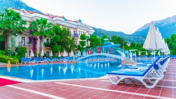 Piscina com cadeiras de descanso azuis, uma ponte e montanhas ao fundo.