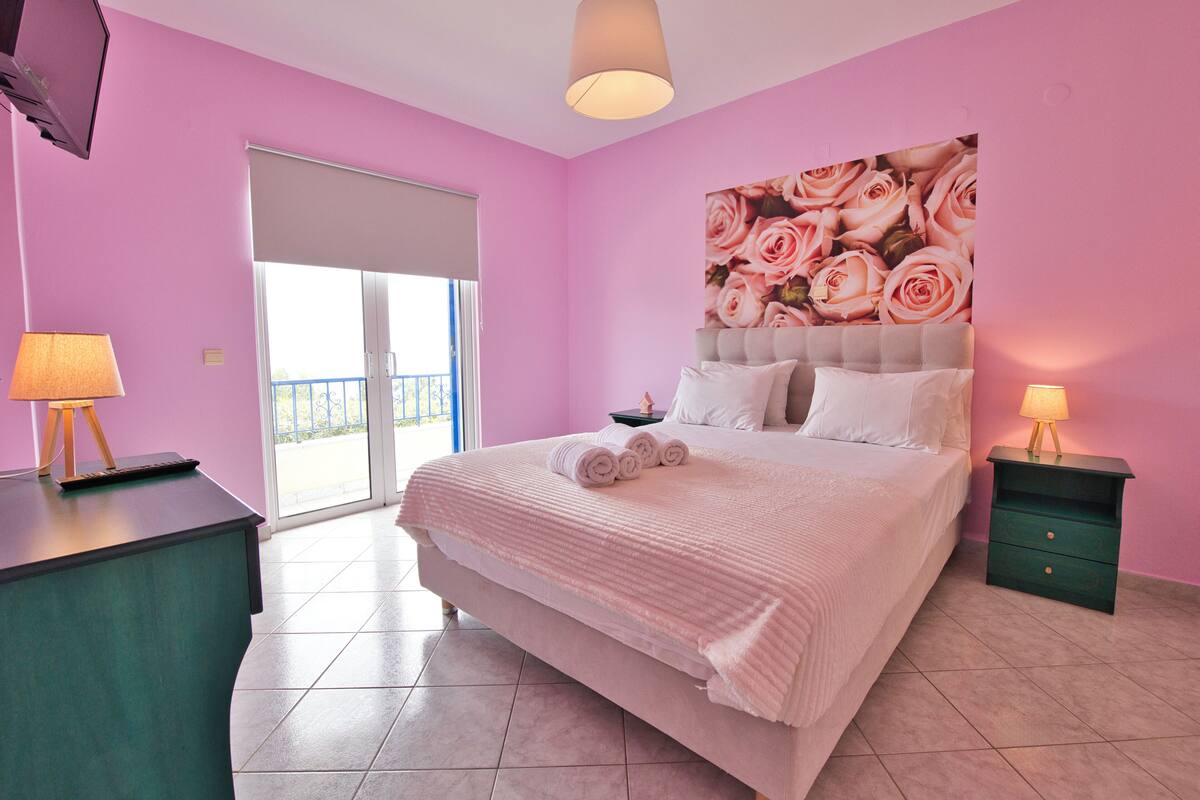 Quarto com uma cama grande, uma porta de correr de vidro para uma varanda, um quadro de rosas na parede e uma mesa de cabeceira verde com uma luminária.