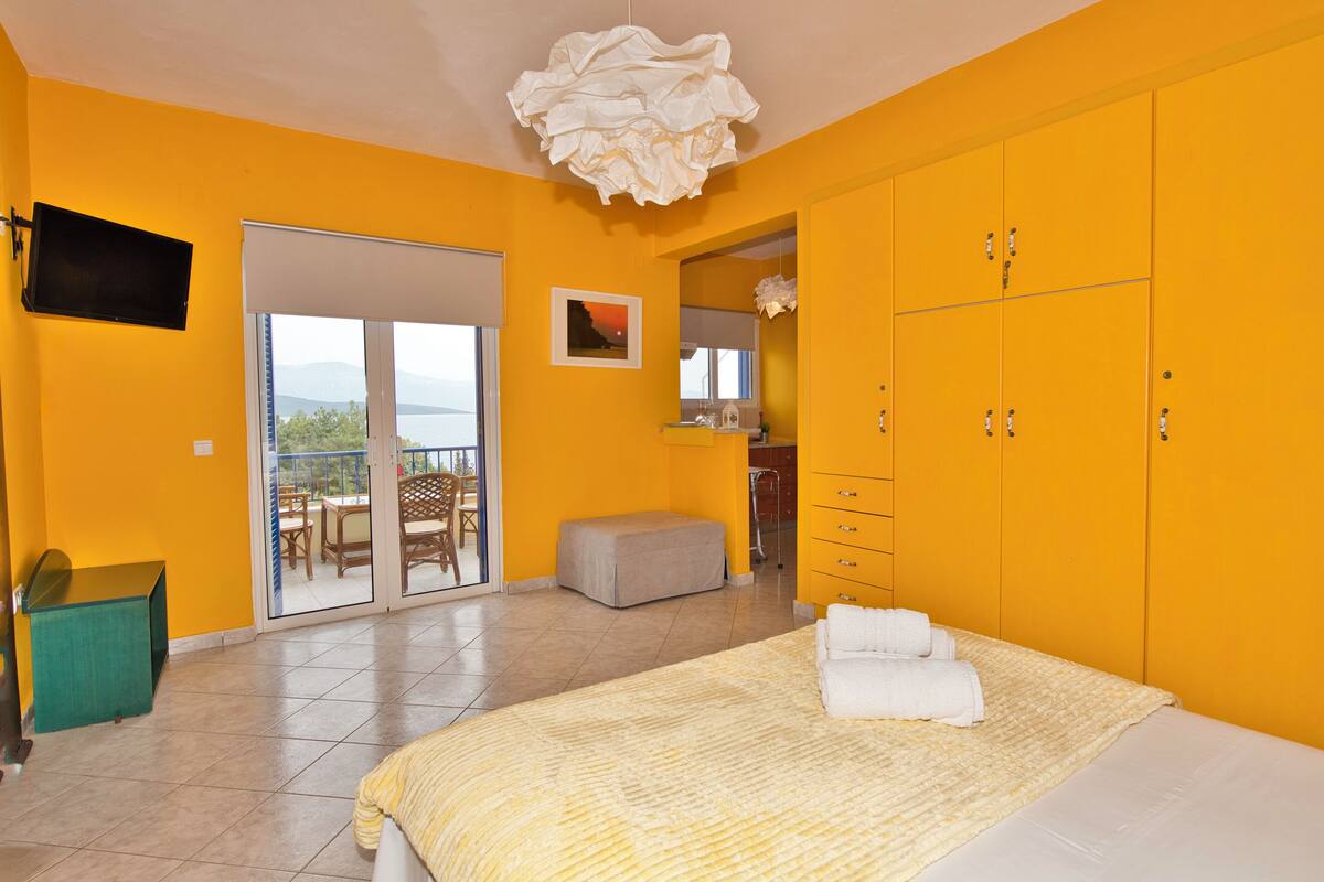 Quarto amarelo e iluminado com cama, televisão e varanda com vista.