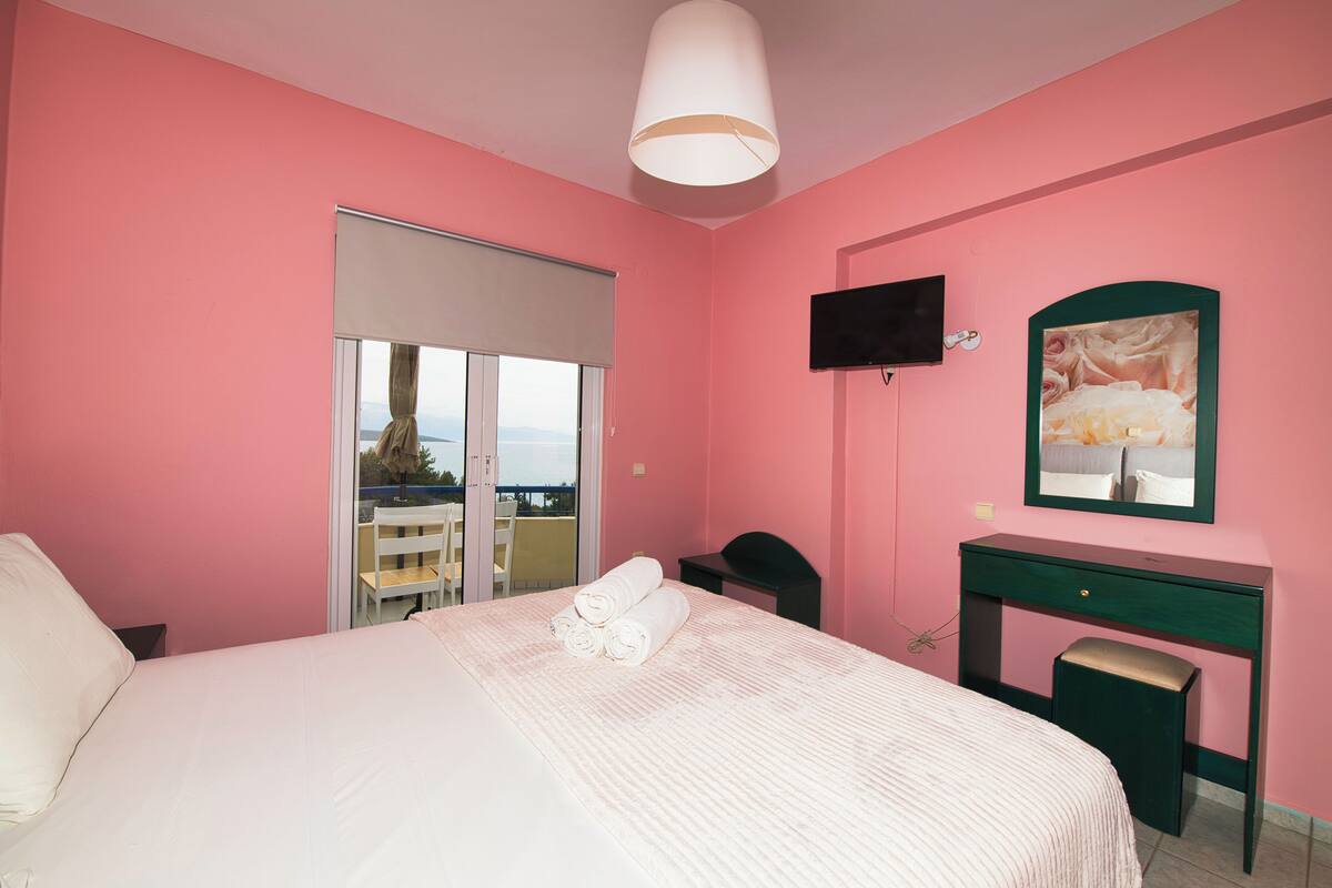 Quarto de hotel em tons de rosa, com cama, escrivaninha, cadeira, televisão e janela com vista.
