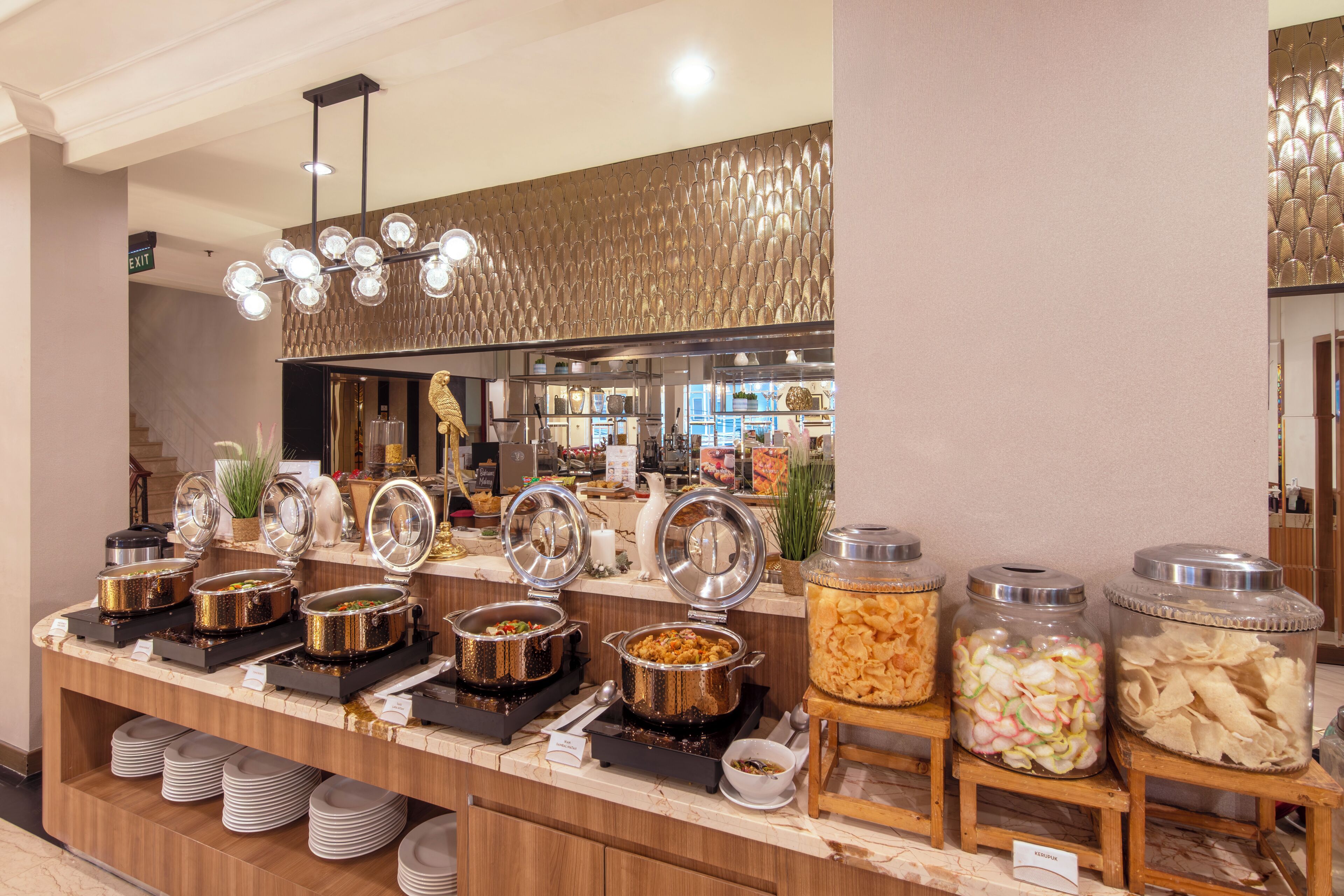Área de buffet com vários recipientes de comida, uma placa indicando a saída e uma parede decorativa com padrão.