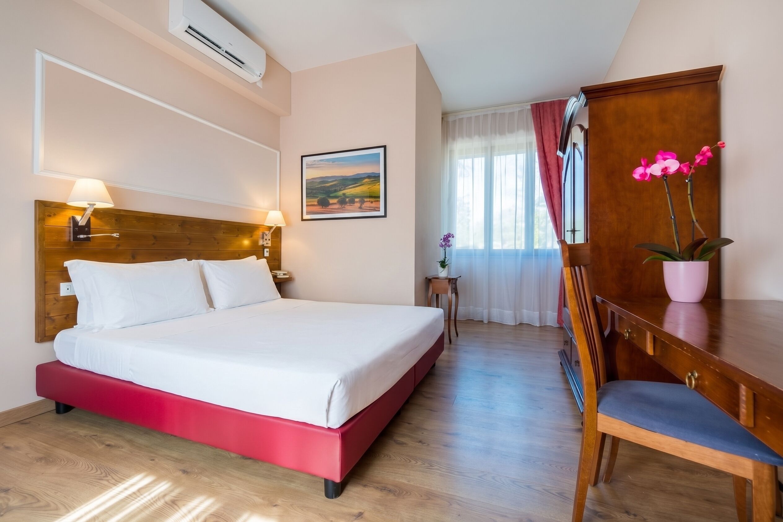 Quarto de hotel com cama, luminárias de cabeceira, escrivaninha, cadeira, ar condicionado, janela com cortinas e um quadro na parede.
