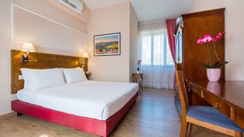 Quarto de hotel com cama, luminárias de cabeceira, escrivaninha, cadeira, ar condicionado, janela com cortinas e um quadro na parede.