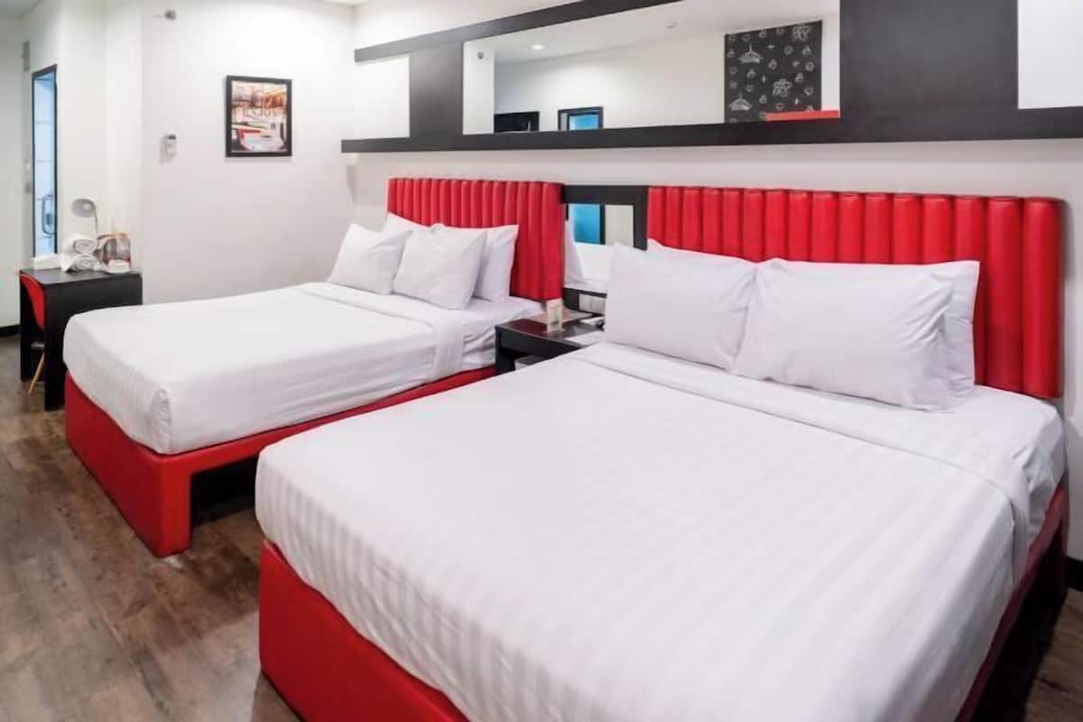 Quarto de hotel com duas camas, uma escrivaninha, uma cadeira e uma parede de destaque vermelha.