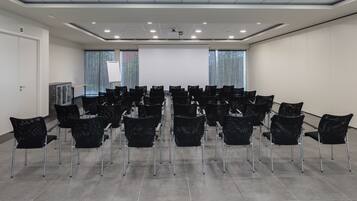 Uma sala de conferências com fileiras de cadeiras pretas, uma tela de projetor e um quadro branco.